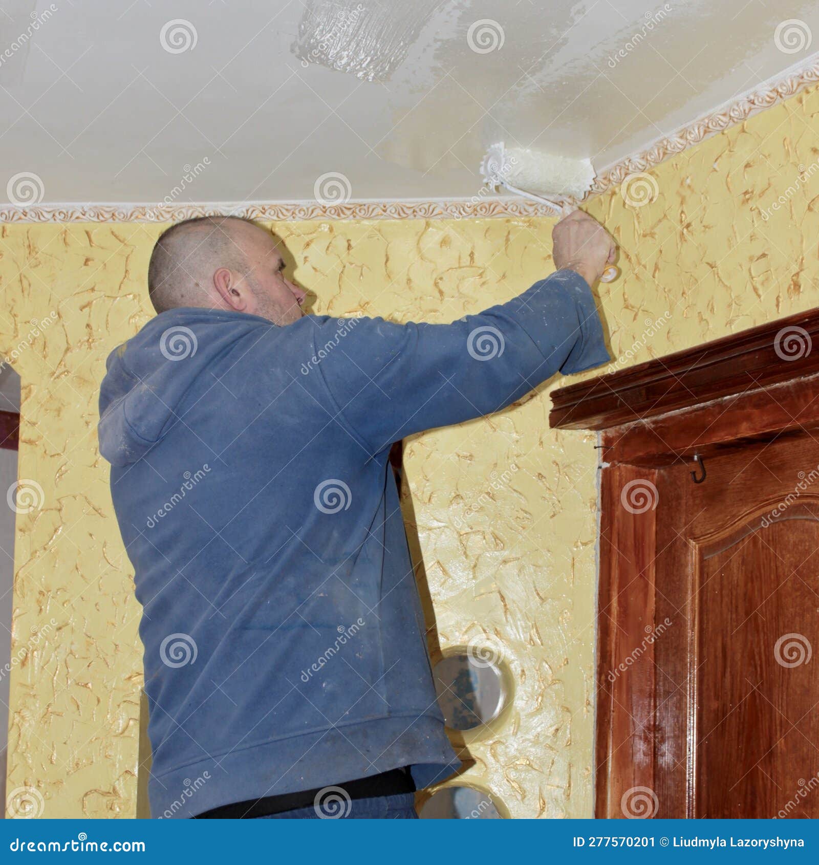 A Male Craftsman in a Blue Suit Paints the Ceiling with a Roller during ...