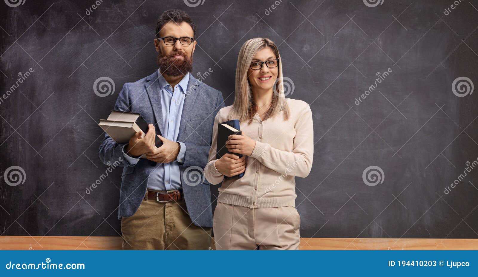 Male And Female Teacher Standing In Front Of A School Blackboard Stock