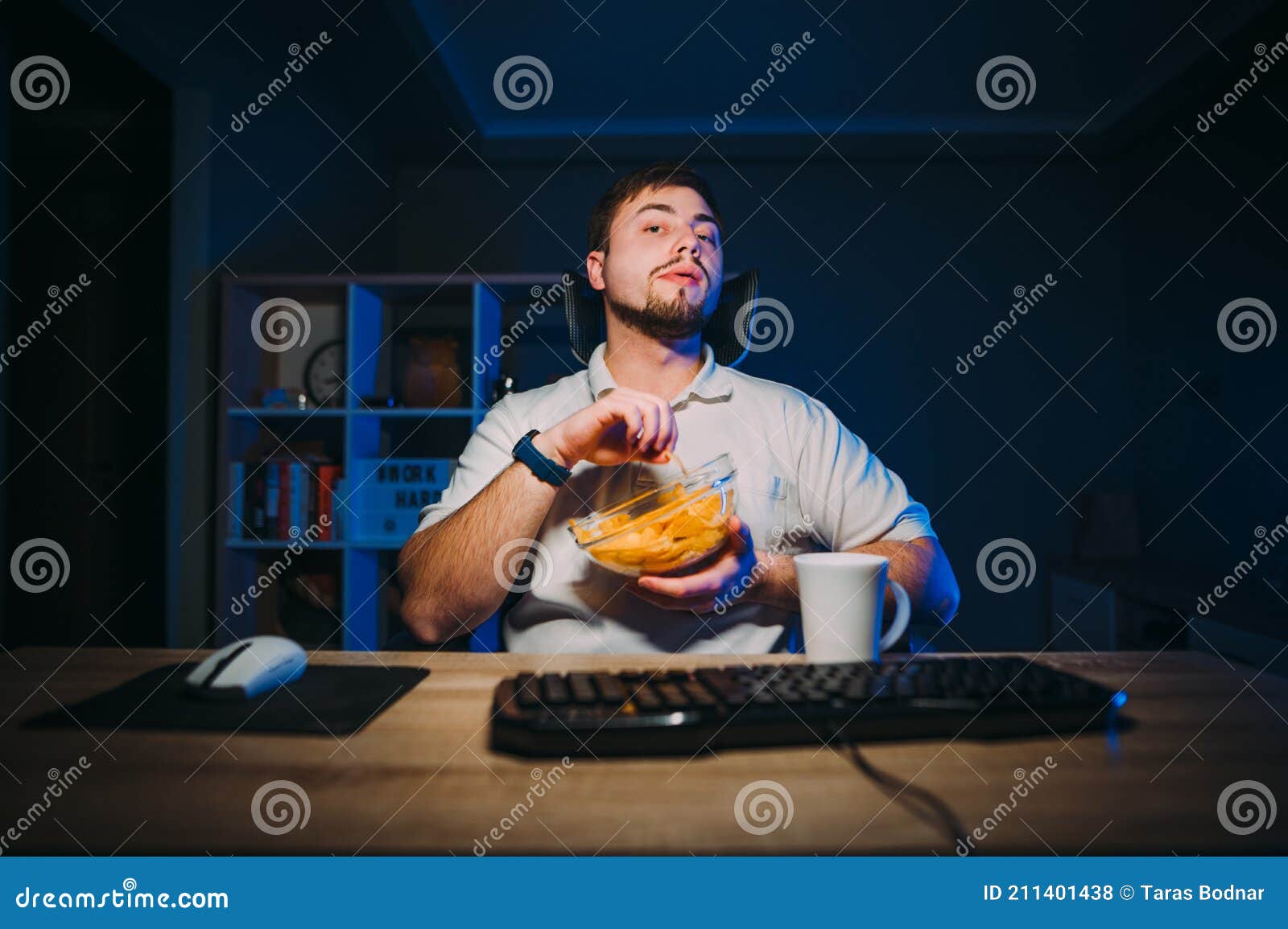 Man with a Beard Works at the Computer at Night and Eats Chips during a ...