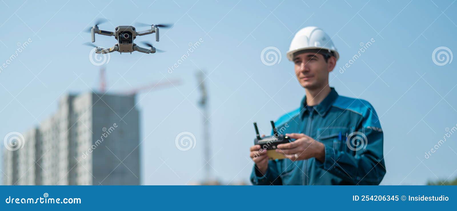 A Man in a Helmet and Overalls Controls a Drone at a Construction Site ...