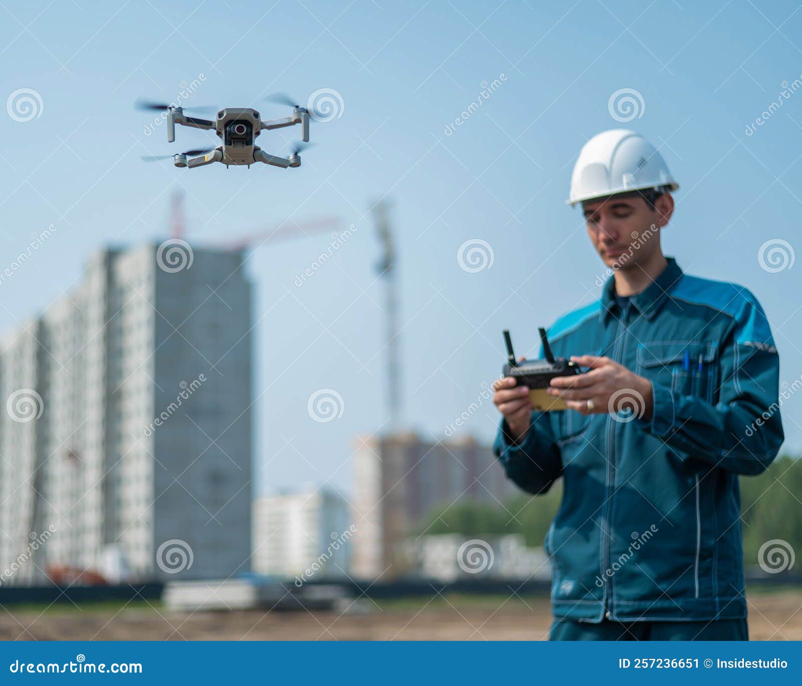 A Man in a Helmet and Overalls Controls a Drone at a Construction Site ...