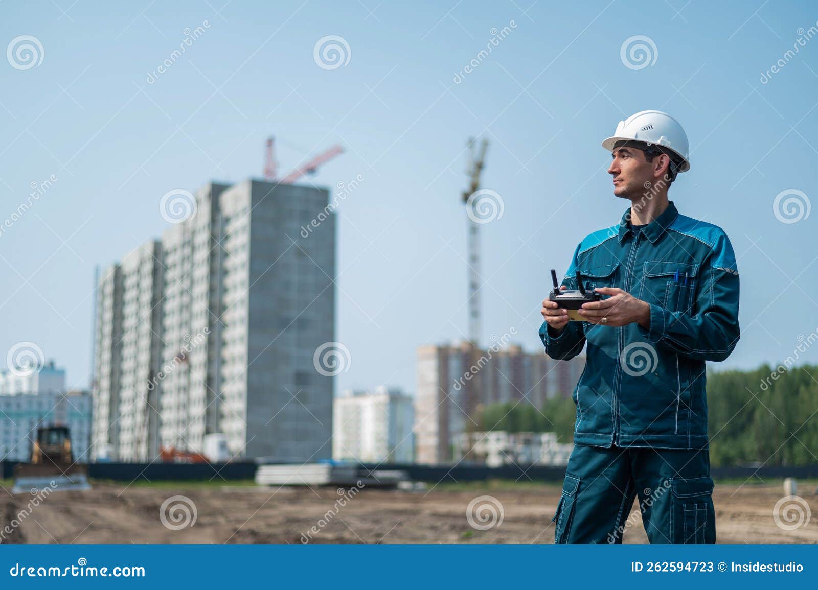 A Man in a Helmet and Overalls Controls a Drone at a Construction Site ...