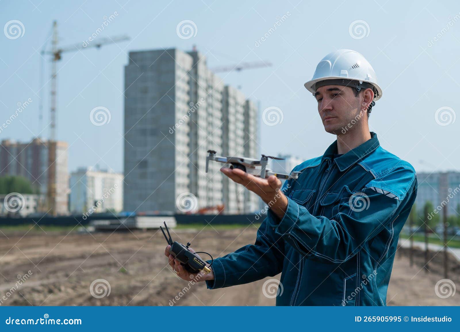 A Man in a Helmet and Overalls Controls a Drone at a Construction Site ...