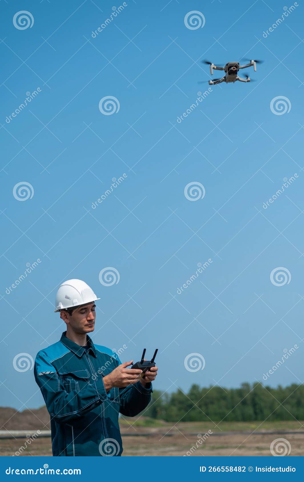 A Man in a Helmet and Overalls Controls a Drone at a Construction Site ...