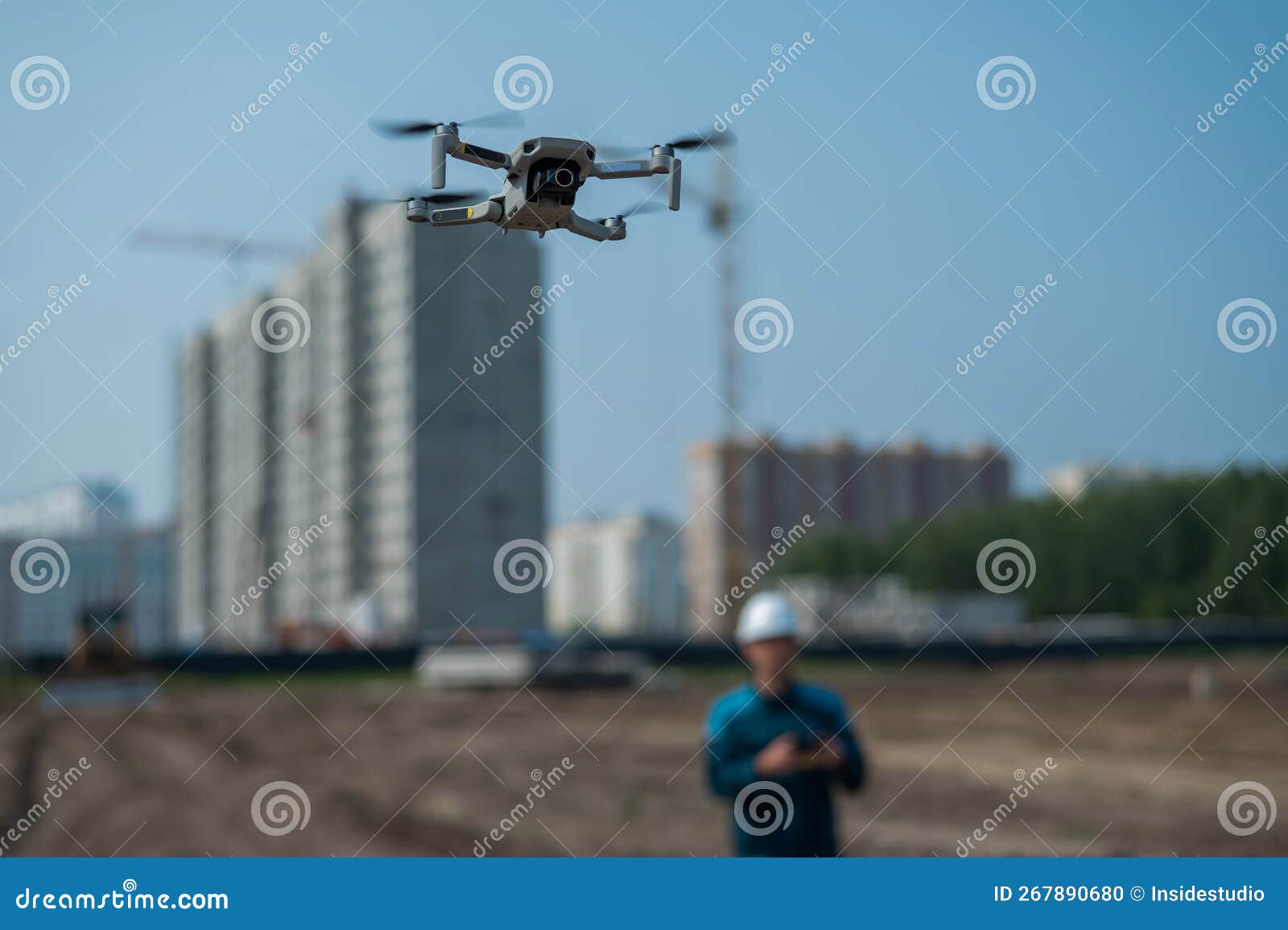 A Man in a Helmet and Overalls Controls a Drone at a Construction Site ...