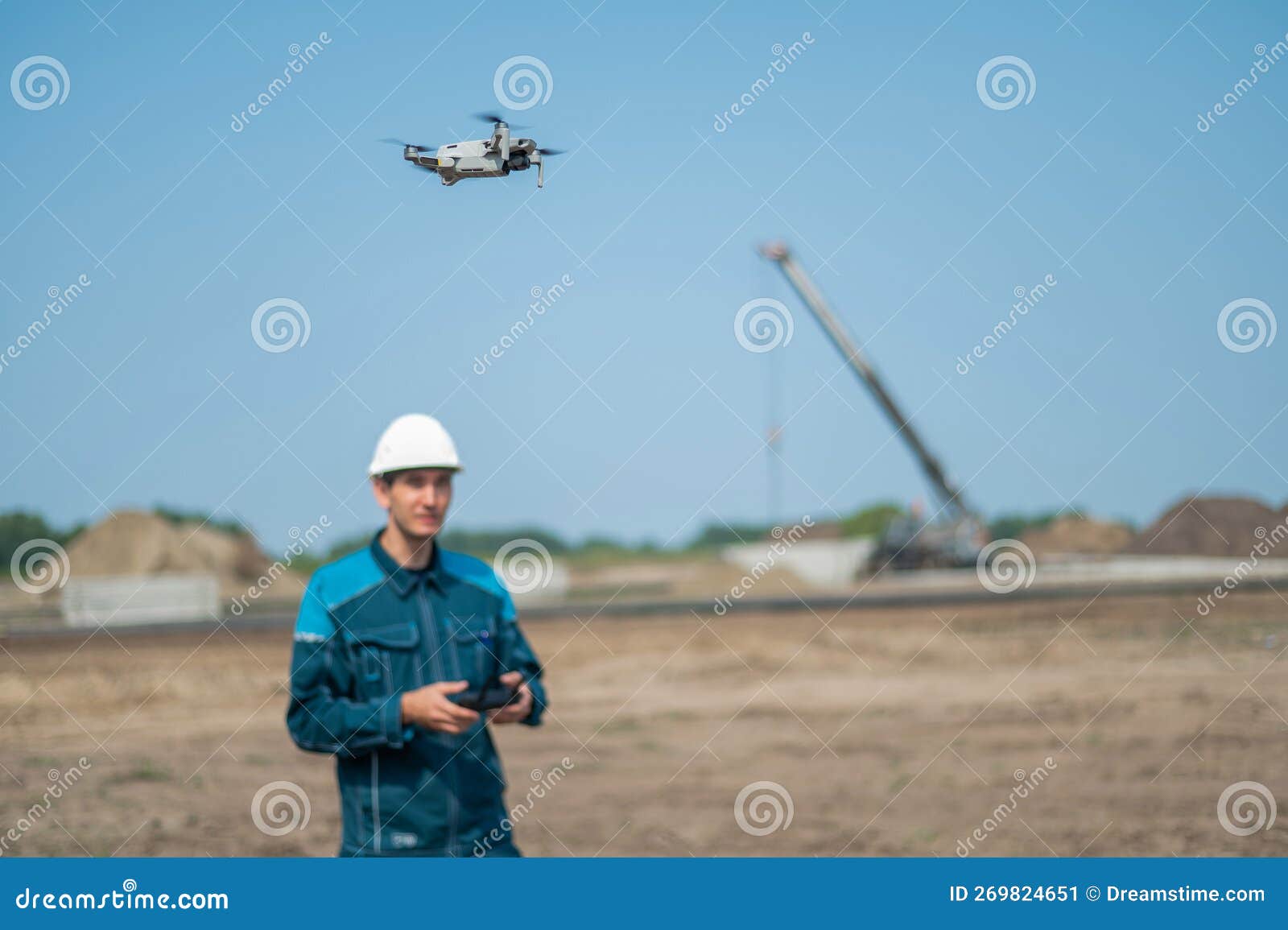A Man in a Helmet and Overalls Controls a Drone at a Construction Site ...