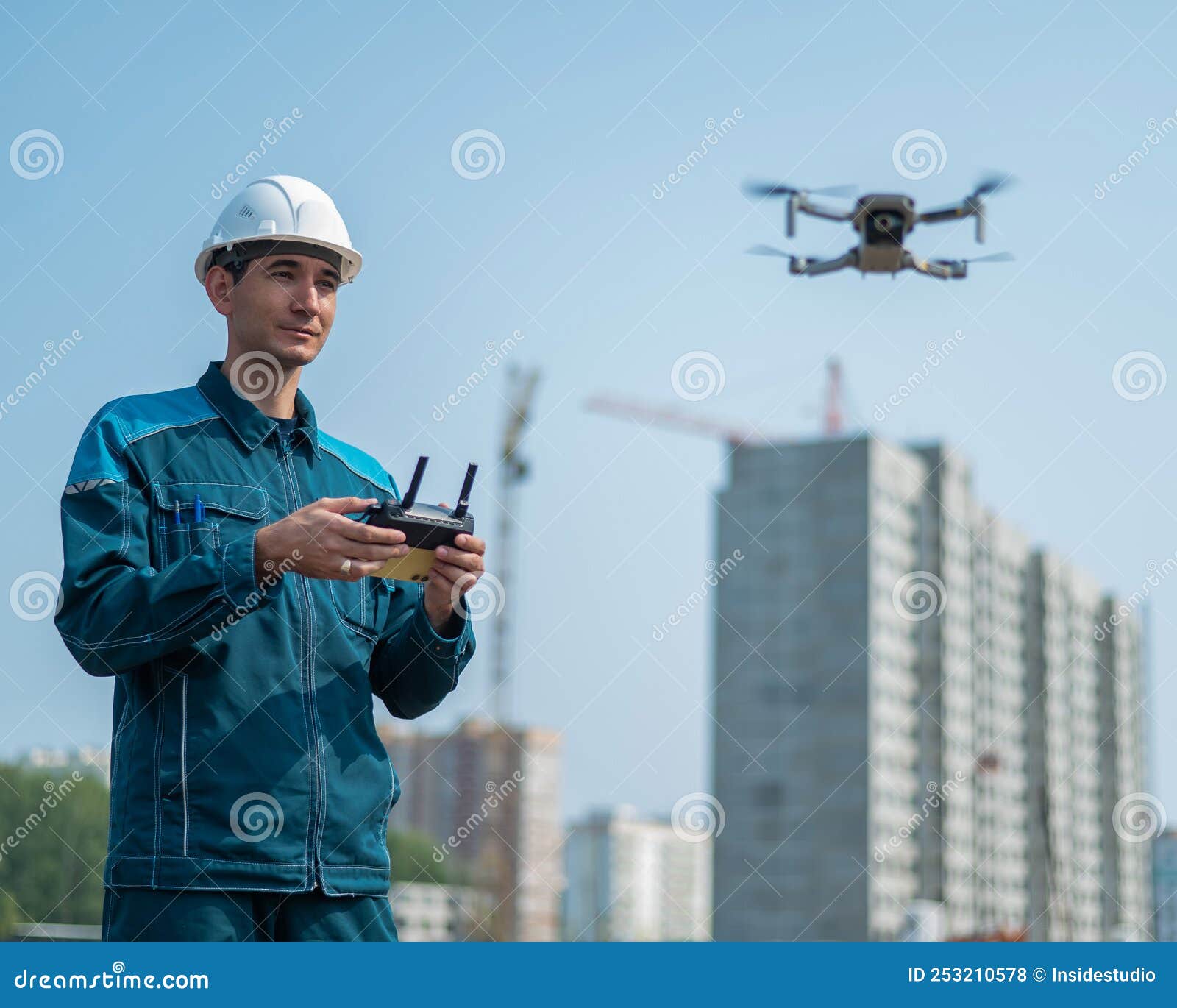 A Man in a Helmet and Overalls Controls a Drone at a Construction Site ...