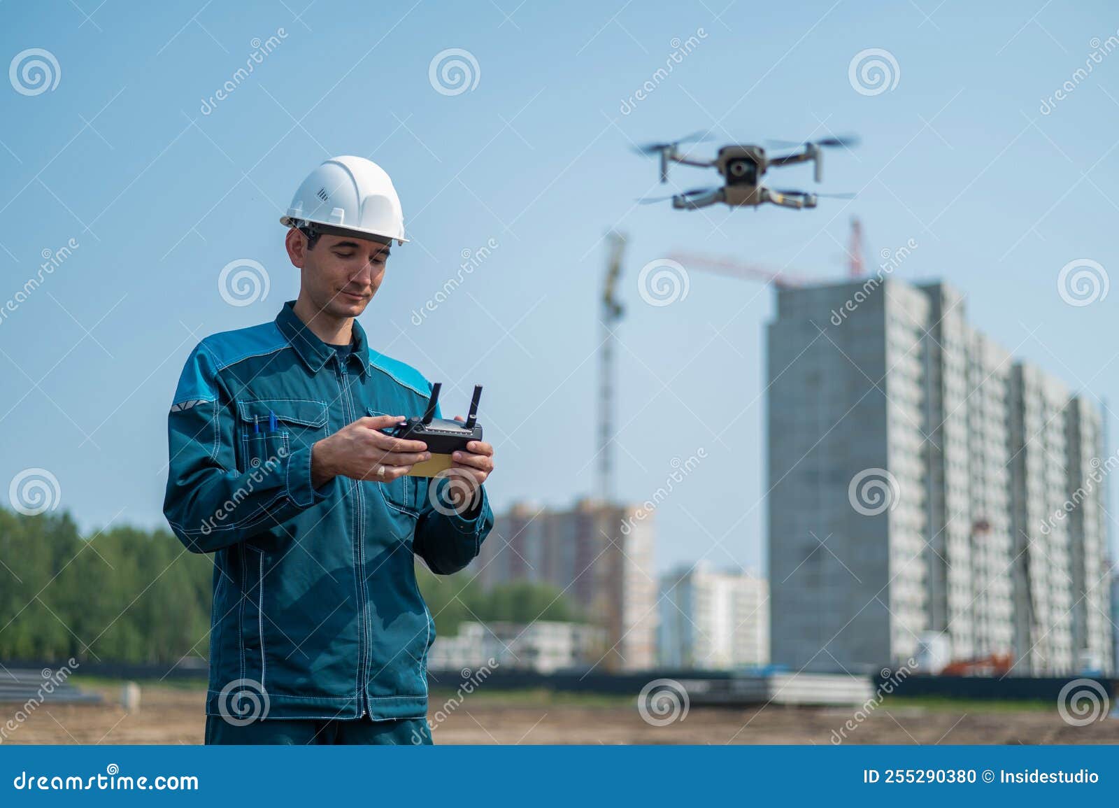 A Man in a Helmet and Overalls Controls a Drone at a Construction Site ...