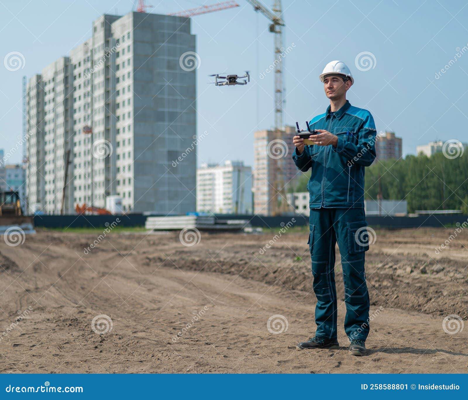A Man in a Helmet and Overalls Controls a Drone at a Construction Site ...