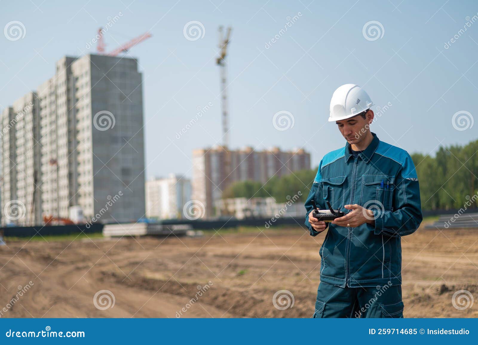 A Man in a Helmet and Overalls Controls a Drone at a Construction Site ...