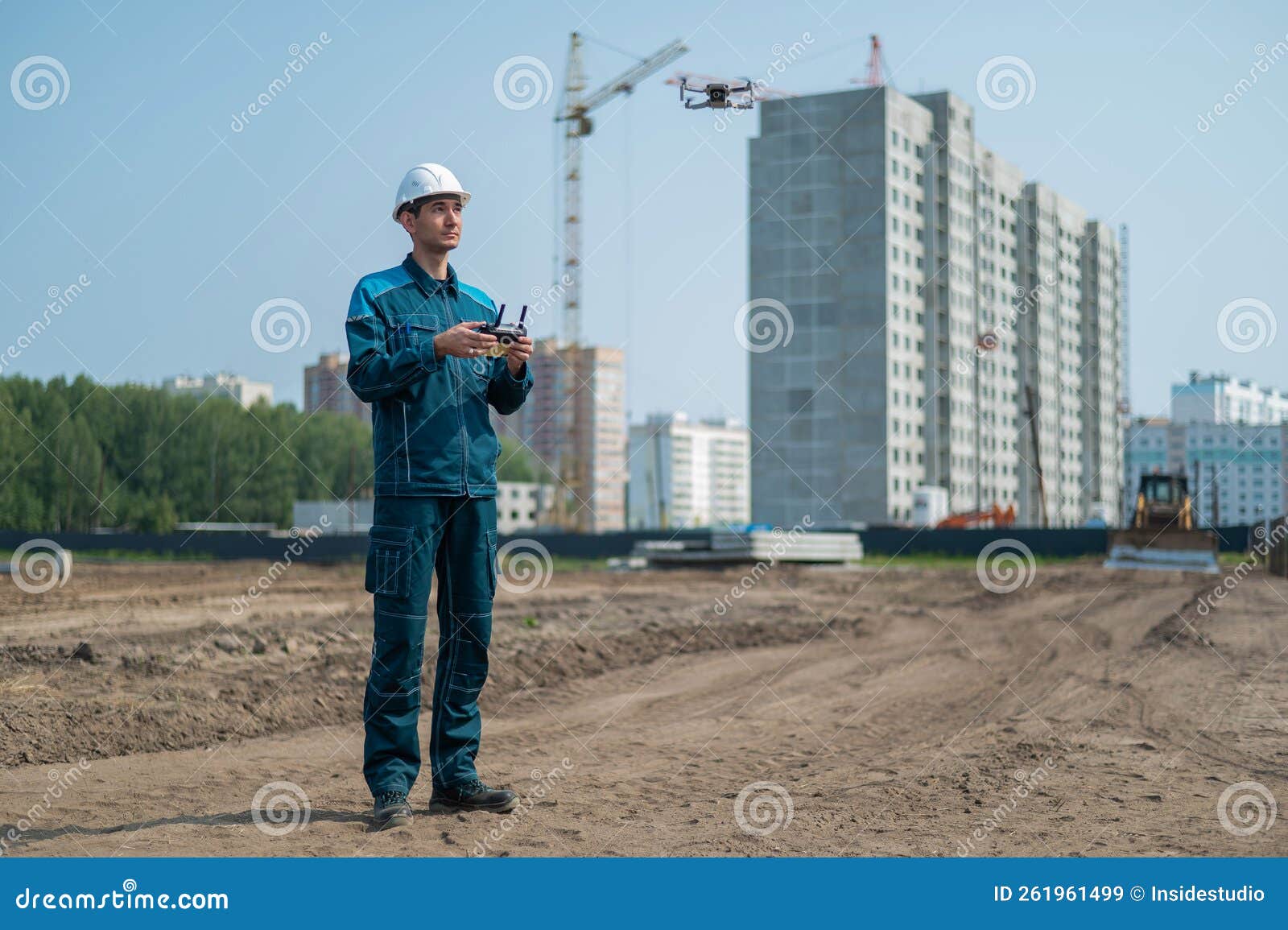 A Man in a Helmet and Overalls Controls a Drone at a Construction Site ...