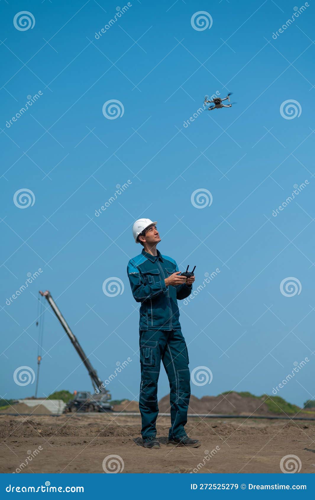 A Man in a Helmet and Overalls Controls a Drone at a Construction Site ...