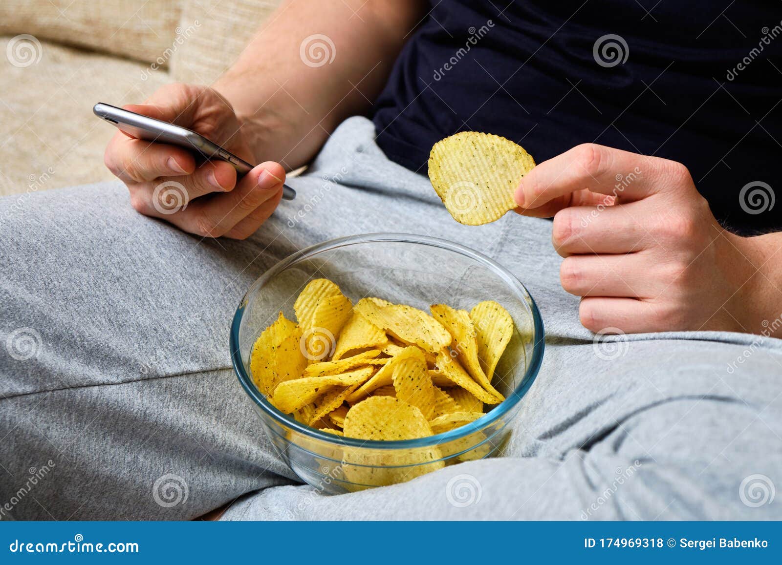 A Man Sits on a Couch Uses a Smartphone and Eats Potato Chips Stock ...