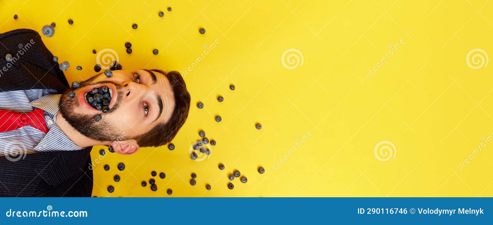 Man In A Suit Lying With Wide Open Mouth Full Of Blueberries On Bright Yellow Background Stock