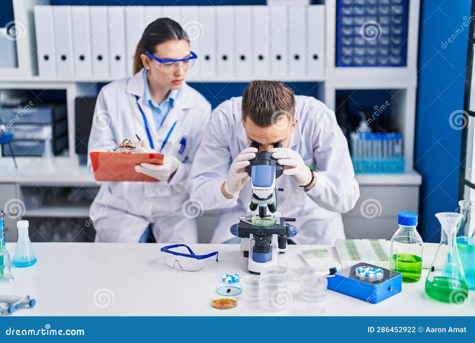 Man and Woman Scientists Using Microscope Write on Clipboard at ...