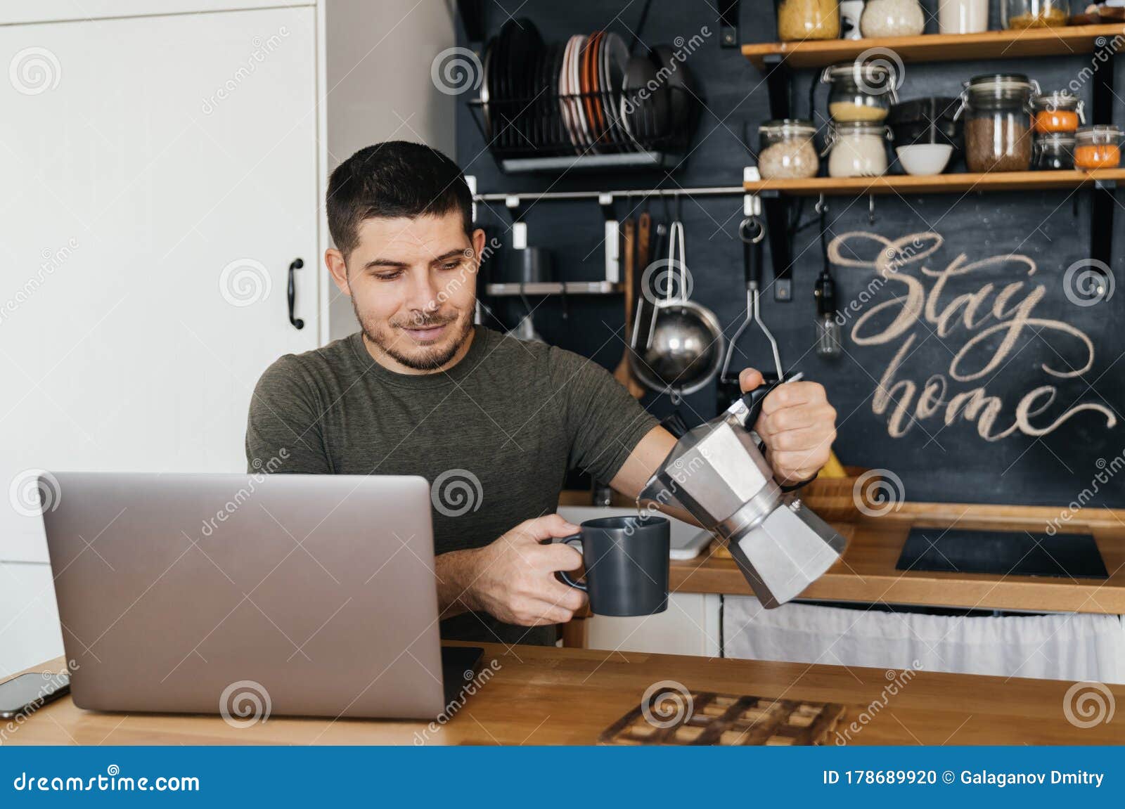 Man Works at a Laptop in the Interior of a Home Kitchen and Pours ...