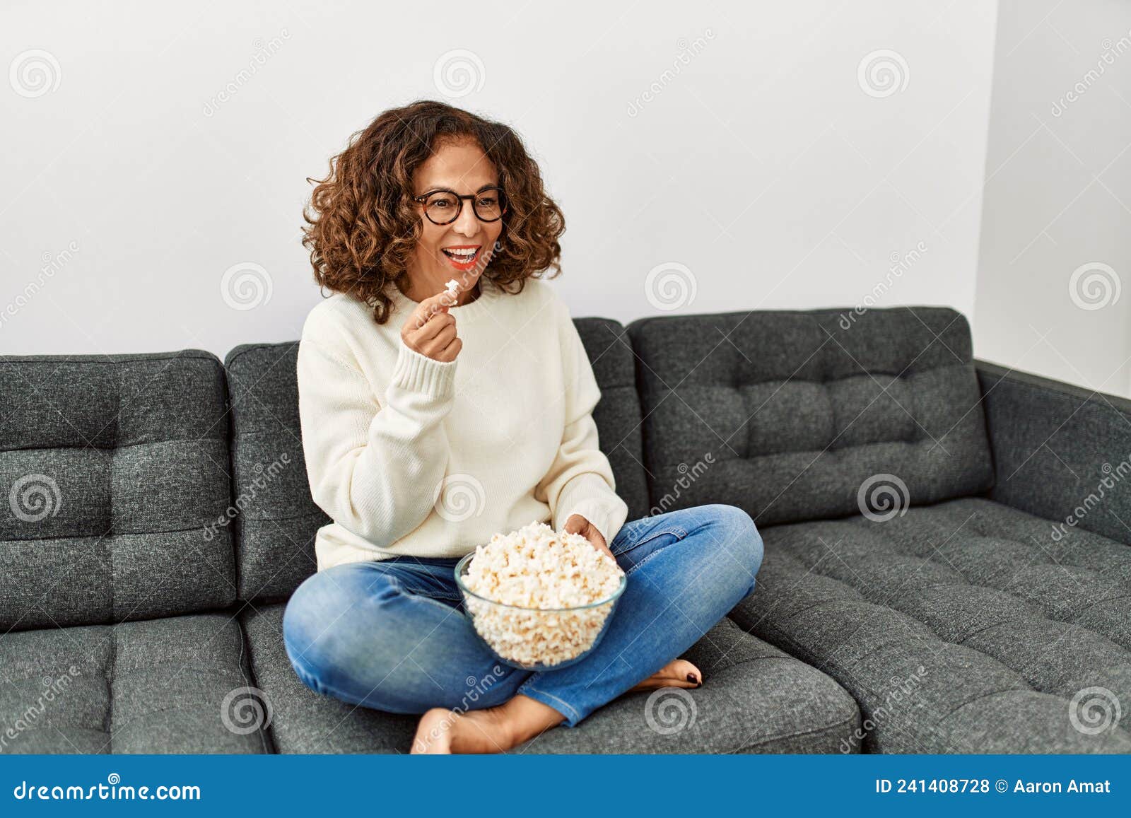 Mature Hispanic Woman Eating Popcorn Watching Tv Sitting On The Sofa At