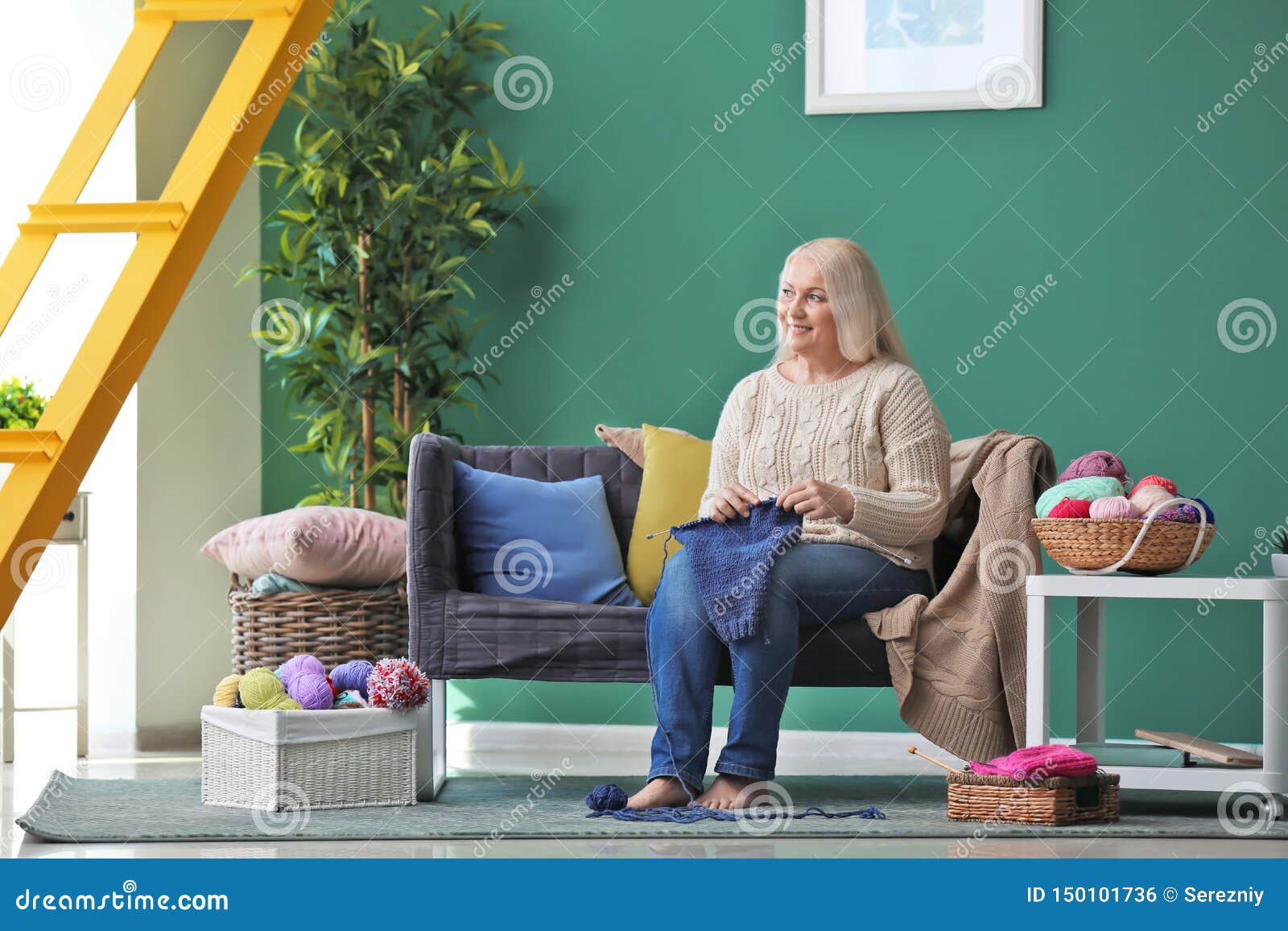 Mature Woman Sitting On Sofa And Knitting Warm Sweater Indoors Stock