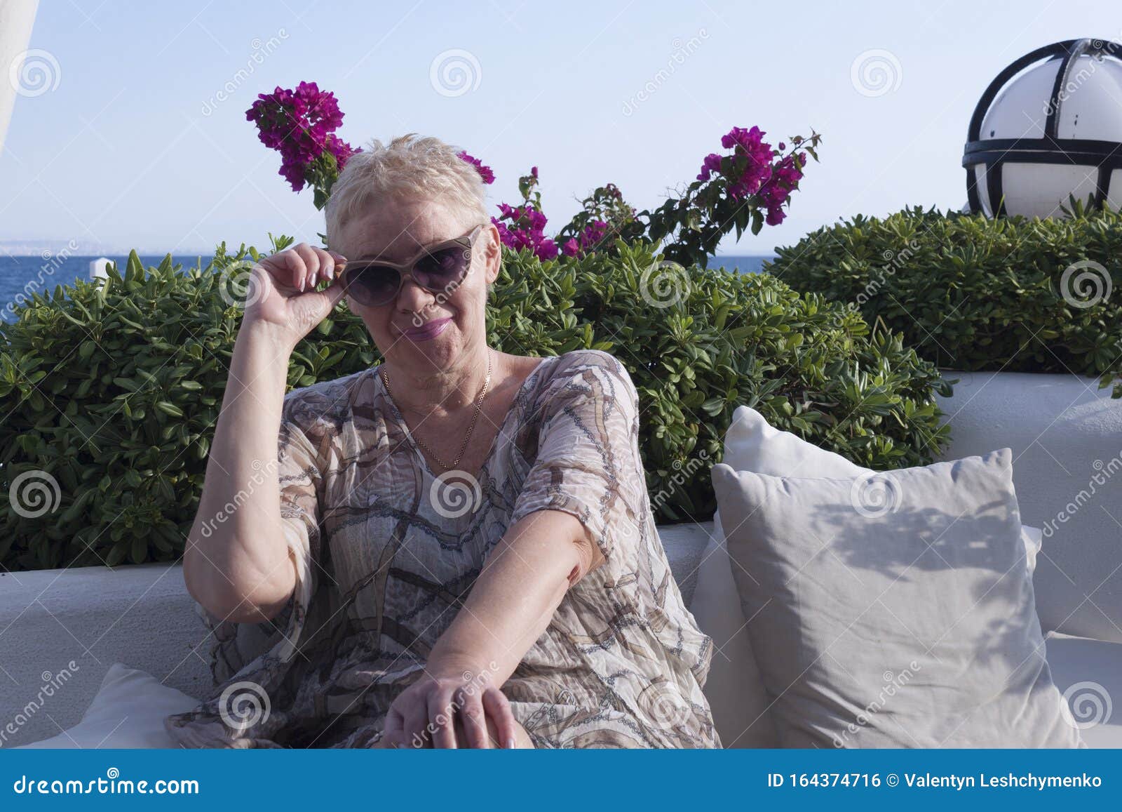 A Mature Woman In Sunglasses Is Smiling While Sitting On A Background