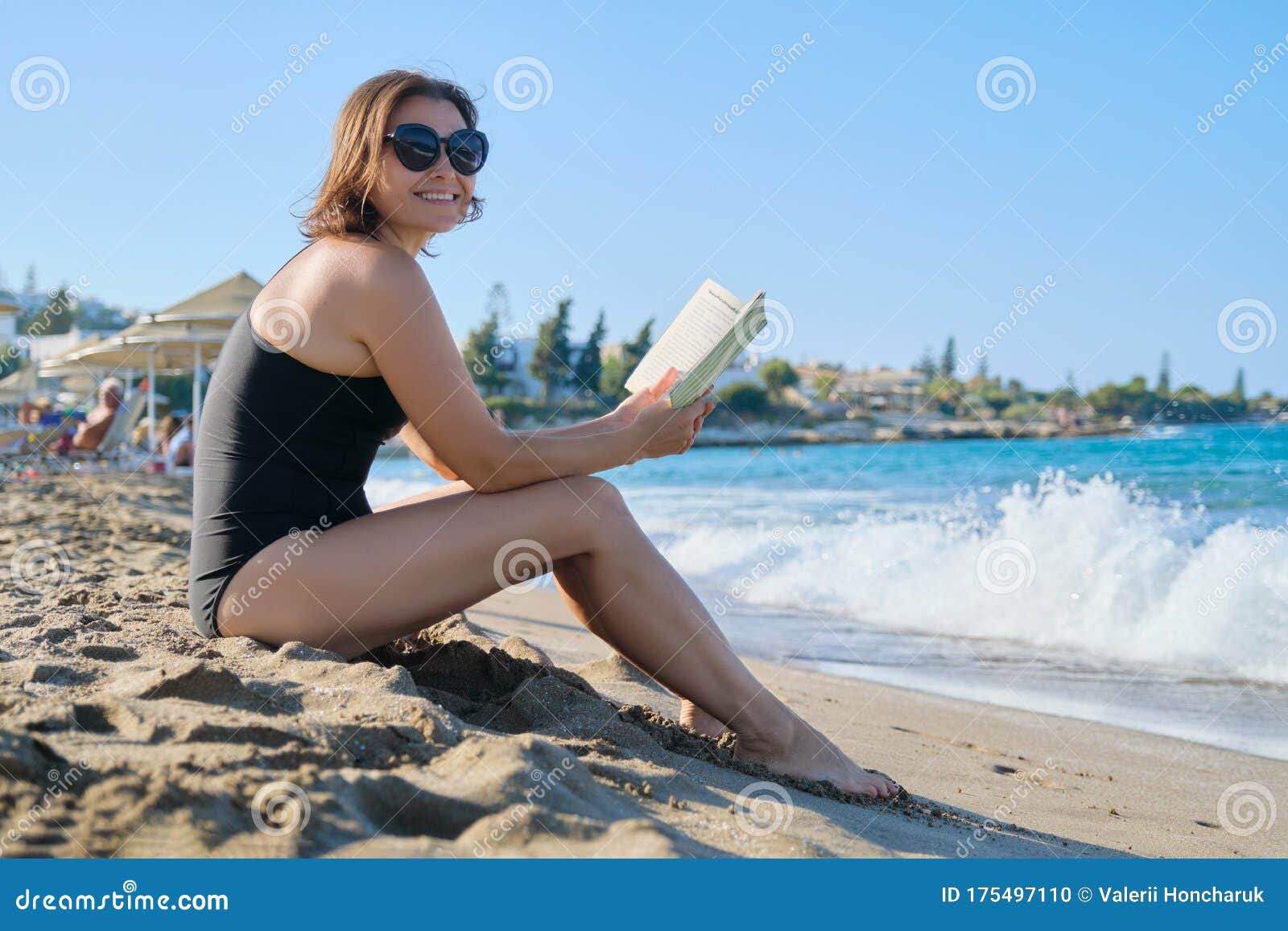 Mature Woman In Sunglasses Swimsuit Reading Book Sitting On Sand By Sea