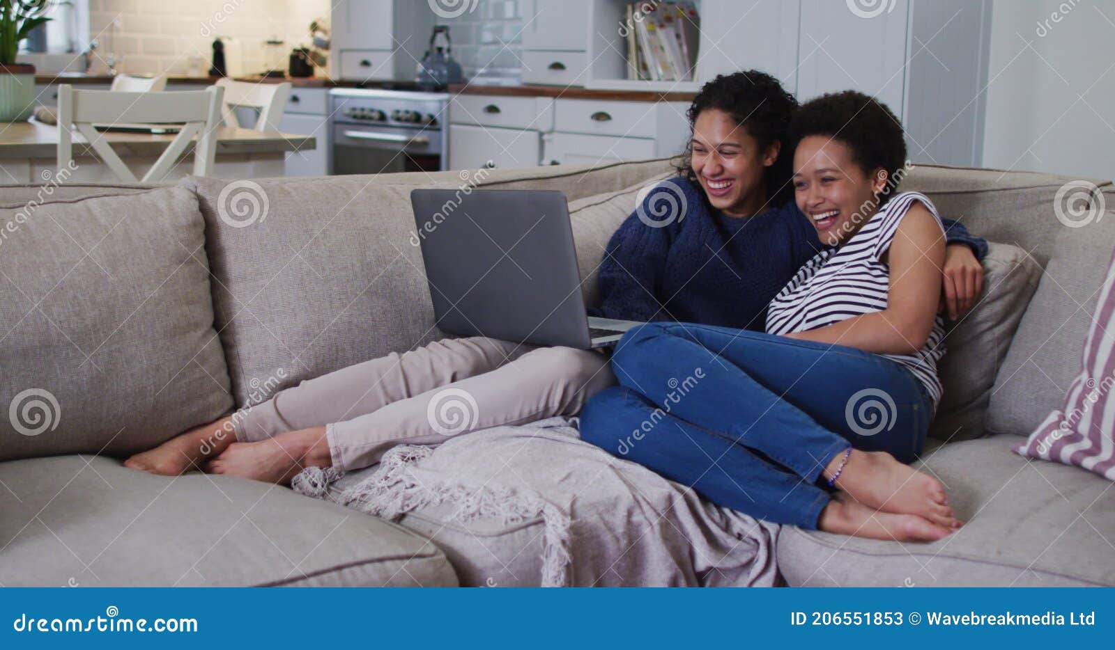 Mixed Race Lesbian Couple Sitting On Couch Embracing And Using Laptop