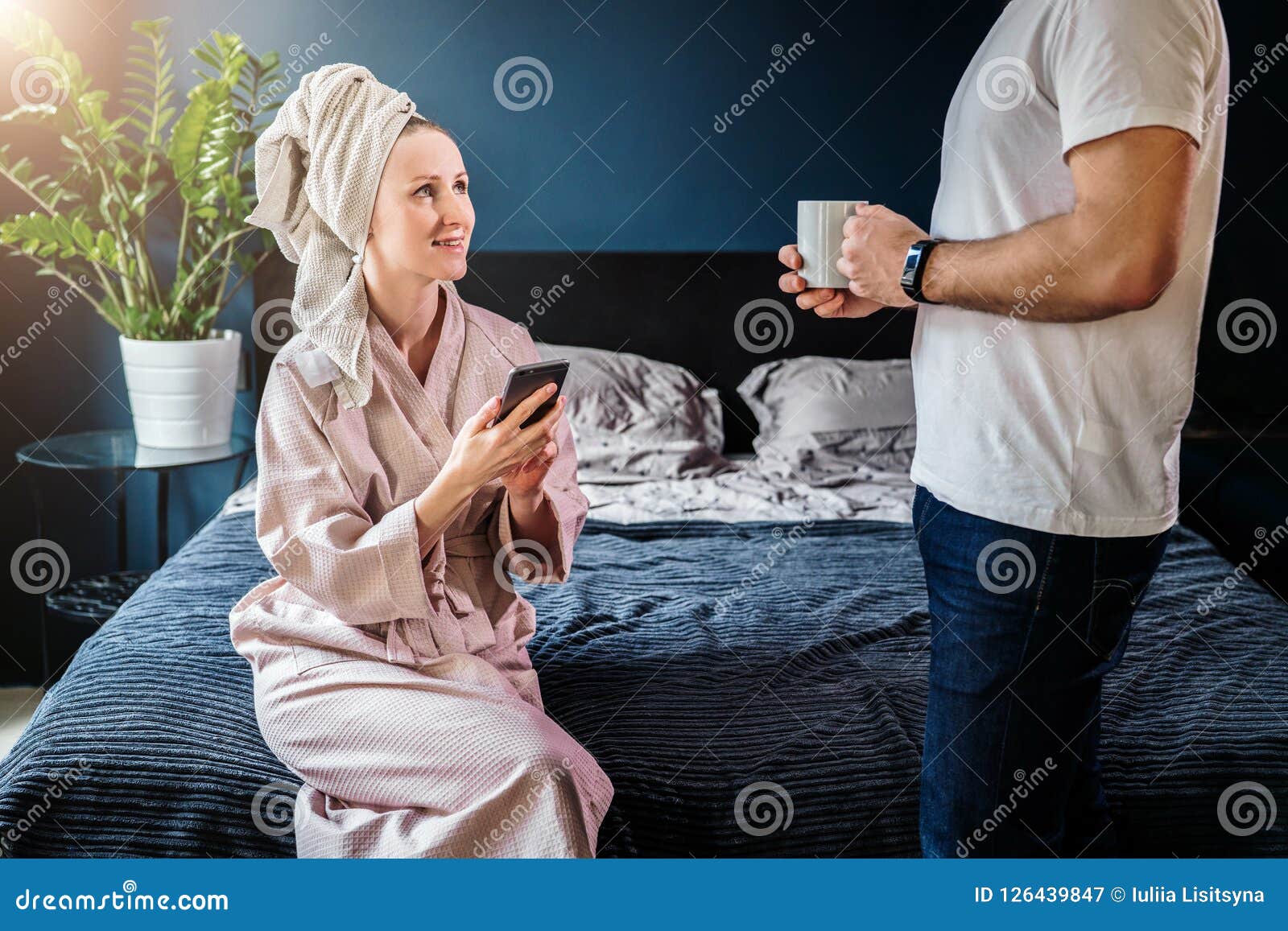 Woman In Bathrobe And Towel On Head Sitting On Bed Holding Smartphone