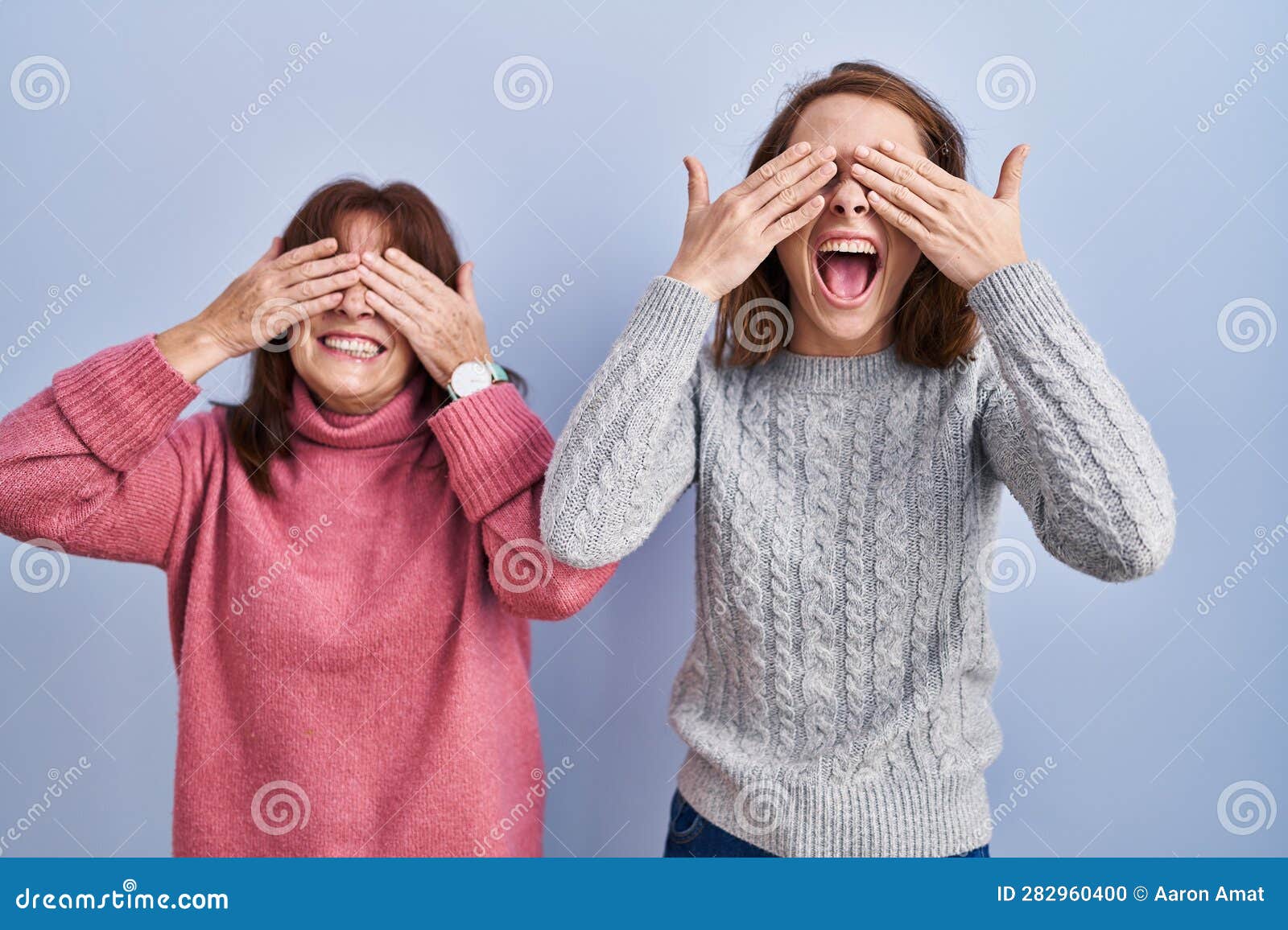 Mother And Daughter Standing Over Blue Background Covering Eyes With
