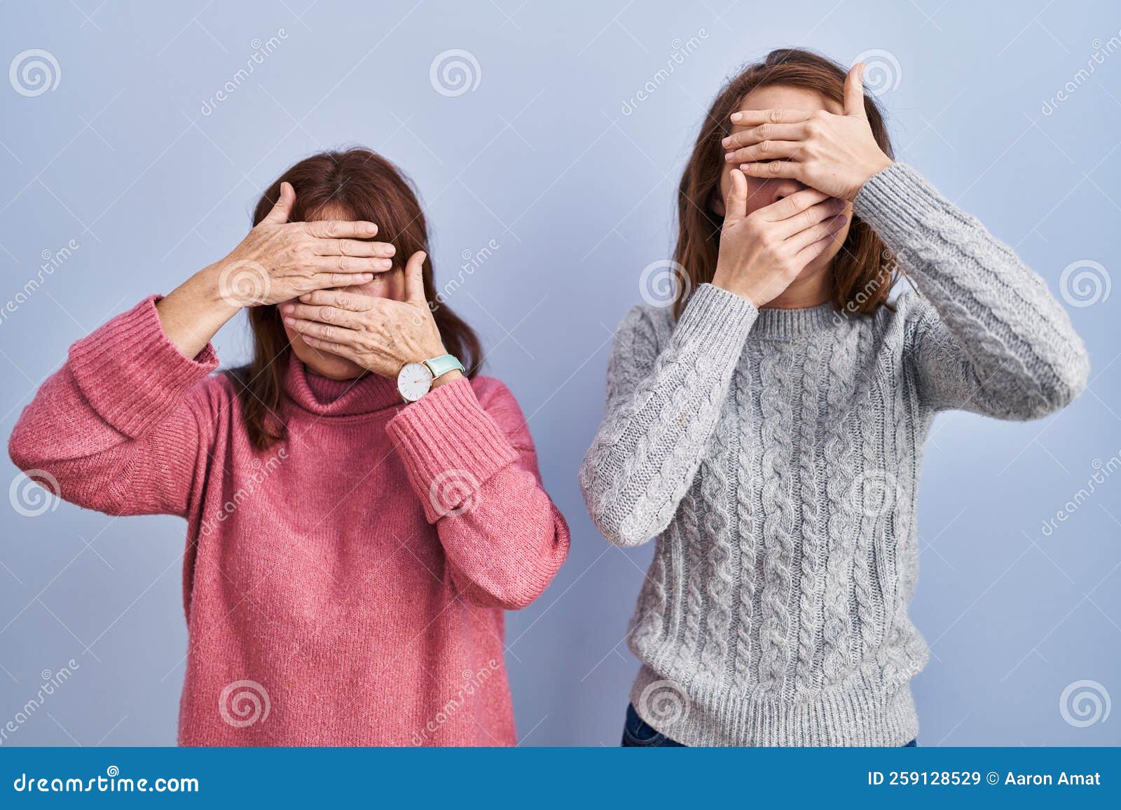 Mother And Daughter Standing Over Blue Background Covering Eyes And