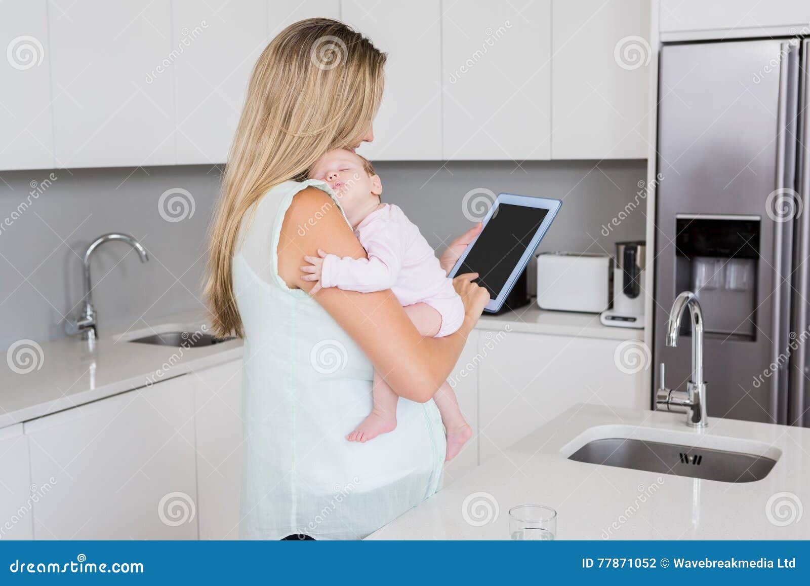 Mother Using Digital Tablet while Carrying Her Baby in Kitchen Stock ...