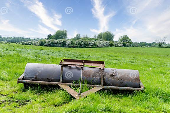 An Old Rusty Farming Roller Lying In A Field With A Copse Of Bushes And Trees In The Far