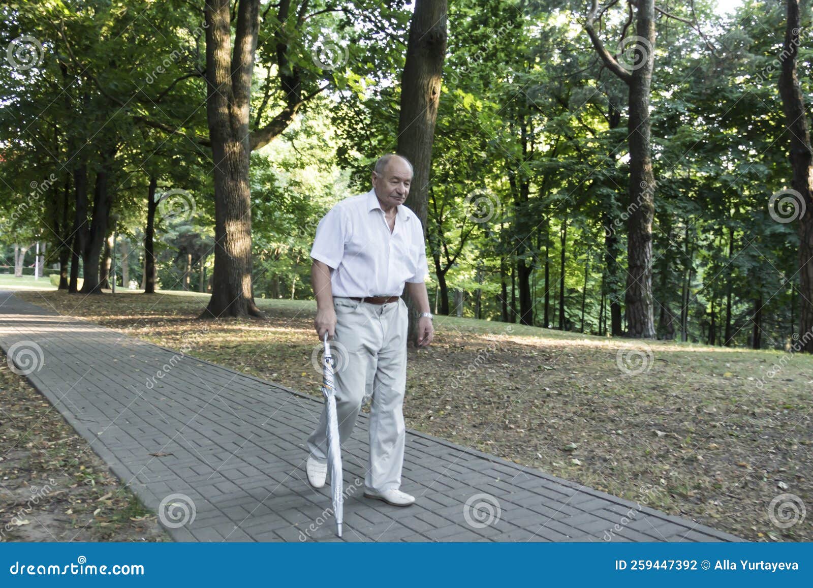 An Old, Stooped Man Walks Sullenly And Alone Along The Path In The Park ...