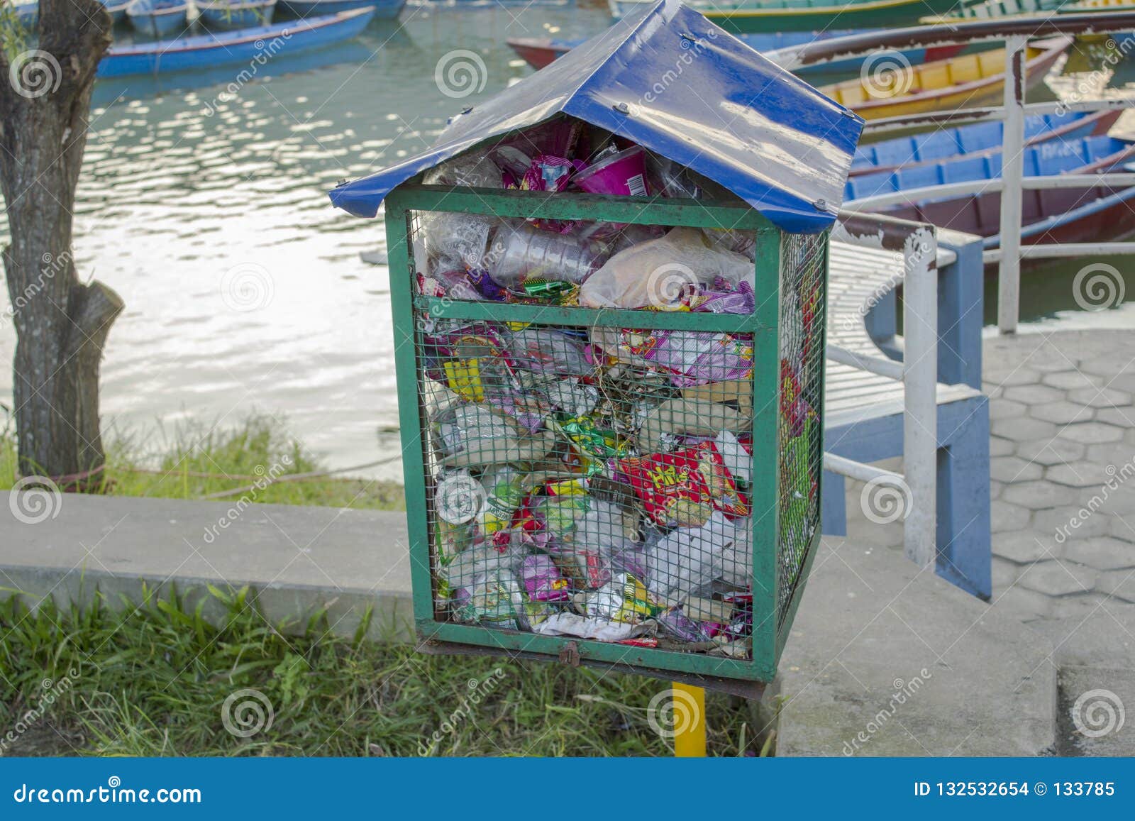 Overflowing Trash Can Near a Bench in a Park by the Lake with Boats ... Overflowing Trash Can Near a Bench in a Park by the Lake with Boats ...