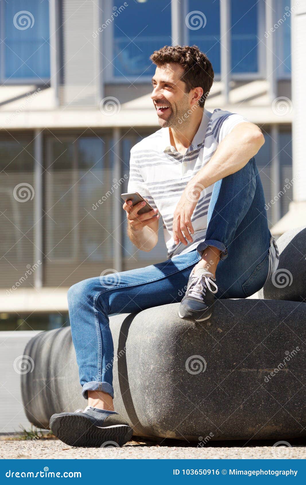 Attractive Young Man Sitting Outdoors With Mobile Phone And Laughing
