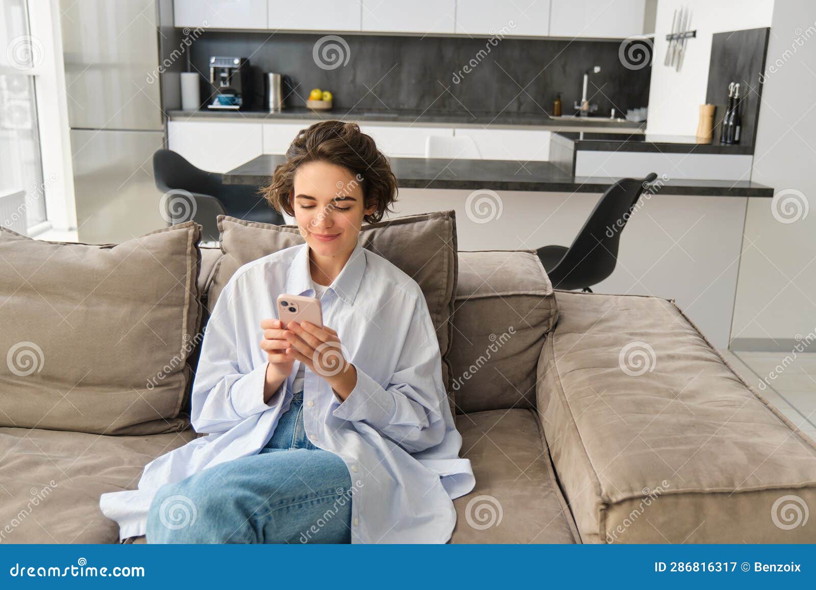 Portrait Of Brunette Woman Sits On Sofa With Smartphone Chats On Phone