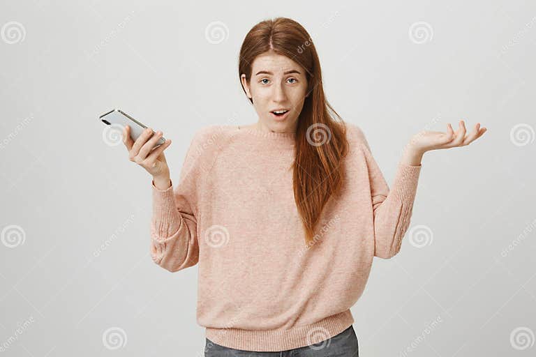 Portrait Of Clueless Puzzled Ginger Girl Shrugging While Holding Smartphone And Talking To