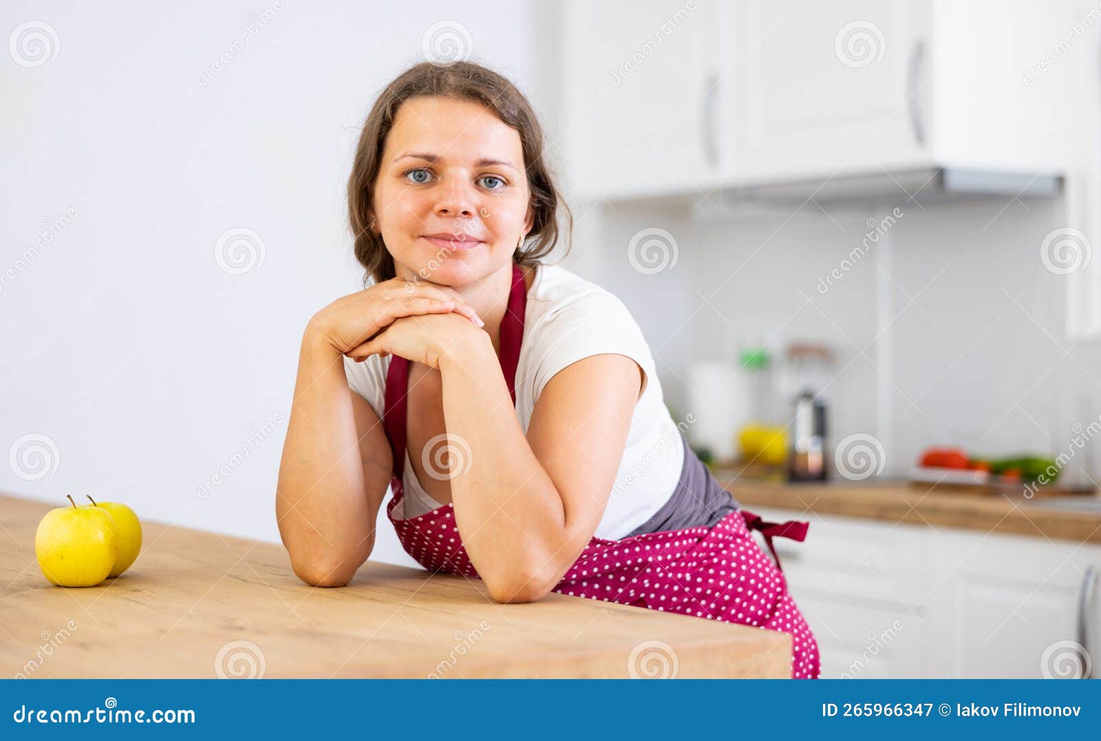 Portrait Of Positive Girl Housewife In Apron Standing At Kitchen Stock