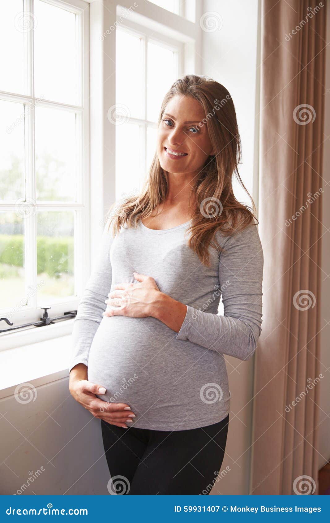 Portrait Of Pregnant Woman Standing By Window Holding Belly Stock Image