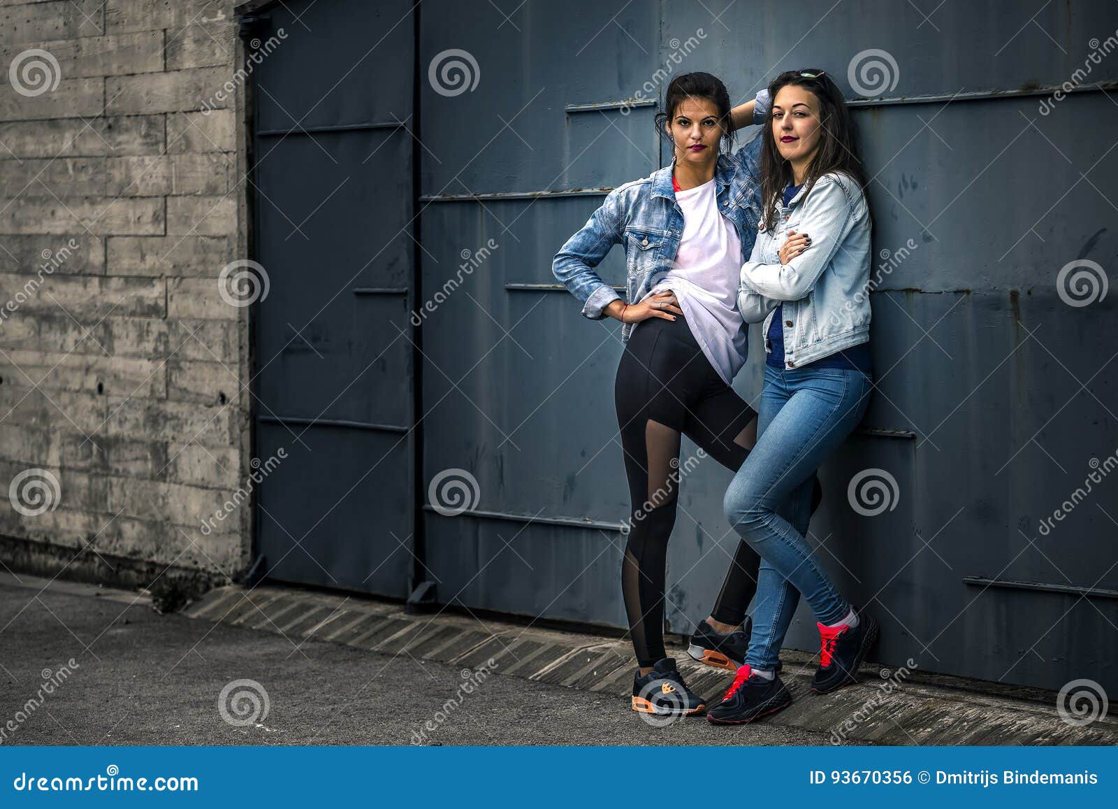 Portrait Of Two Young And Attractive Women Standing Next To The Wall
