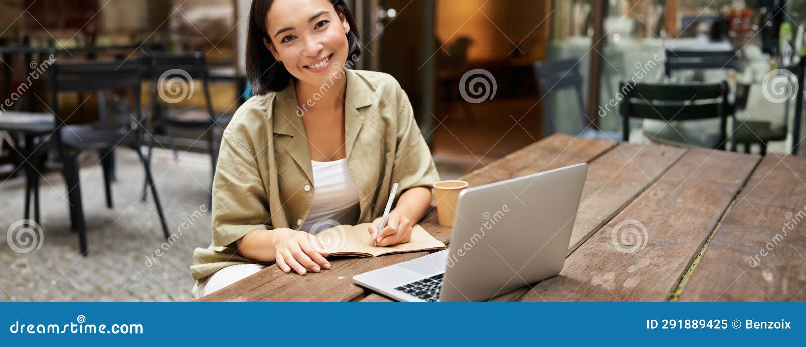 Portrait Of Young Woman Sitting In Cafe With Laptop Making Notes