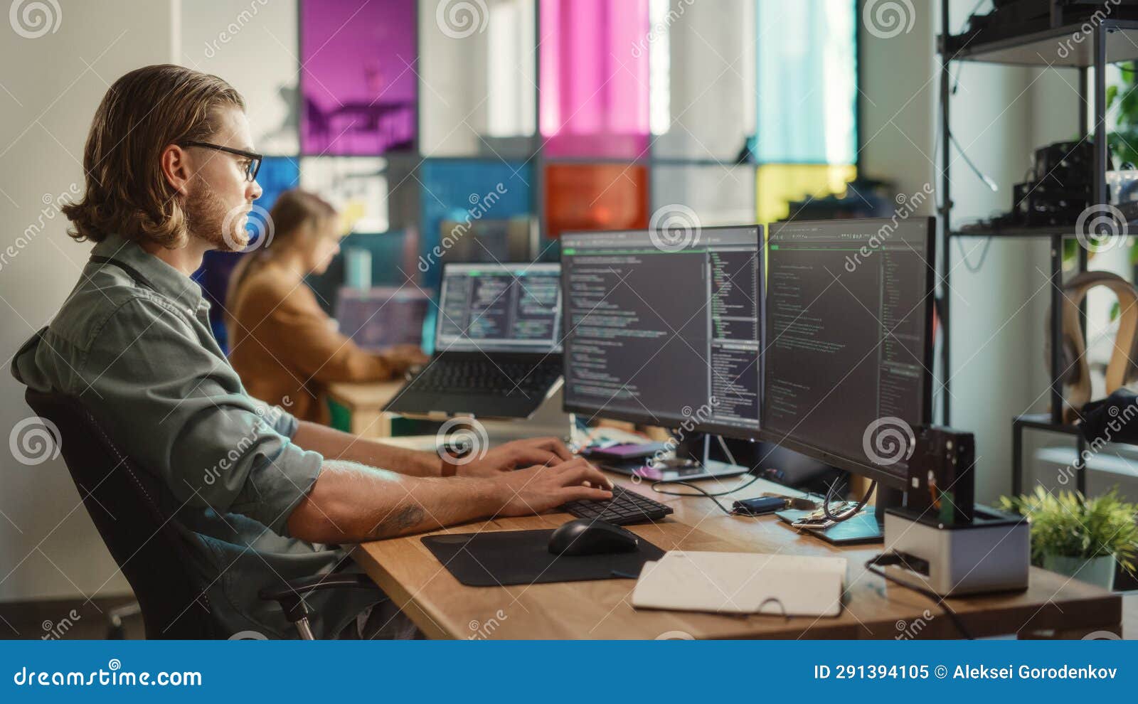 Professional Male Developer Writing Code on Desktop Computer with Two Displays and a Laptop in ...