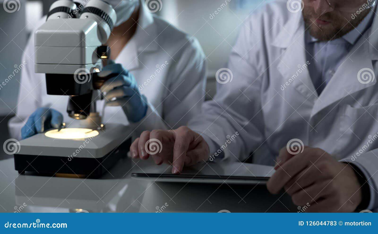 Researcher Studying Sample Under Microscope, Assistant Making Records ...