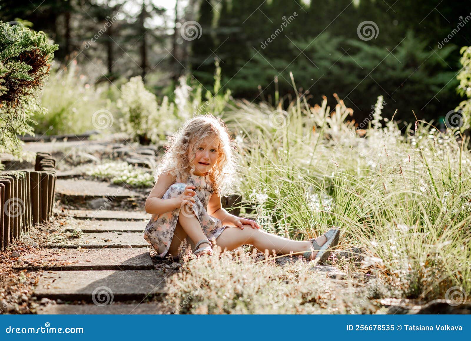 Sad Upset Crying Little Blonde Curly Girl In Dress Sitting On Wood Road In Grass Near Meadow