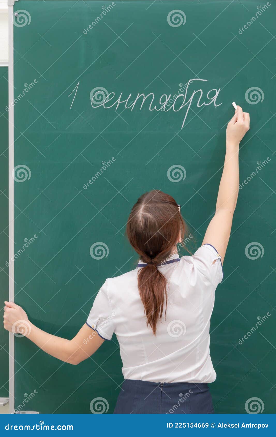 School Teacher Young Woman In A White Blouse In The Classroom On A Green Blackboard Writes With