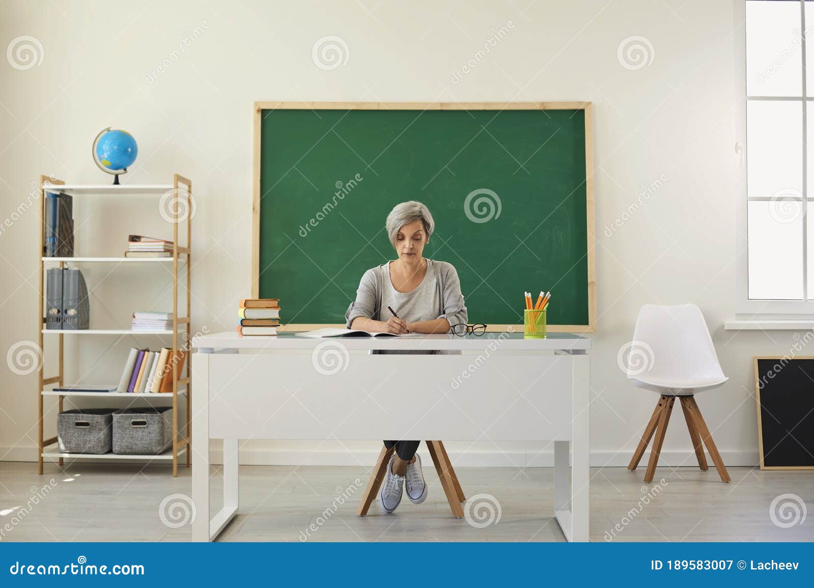 Serious Mature Teacher Sitting At Desk And Writing In Classroom Female