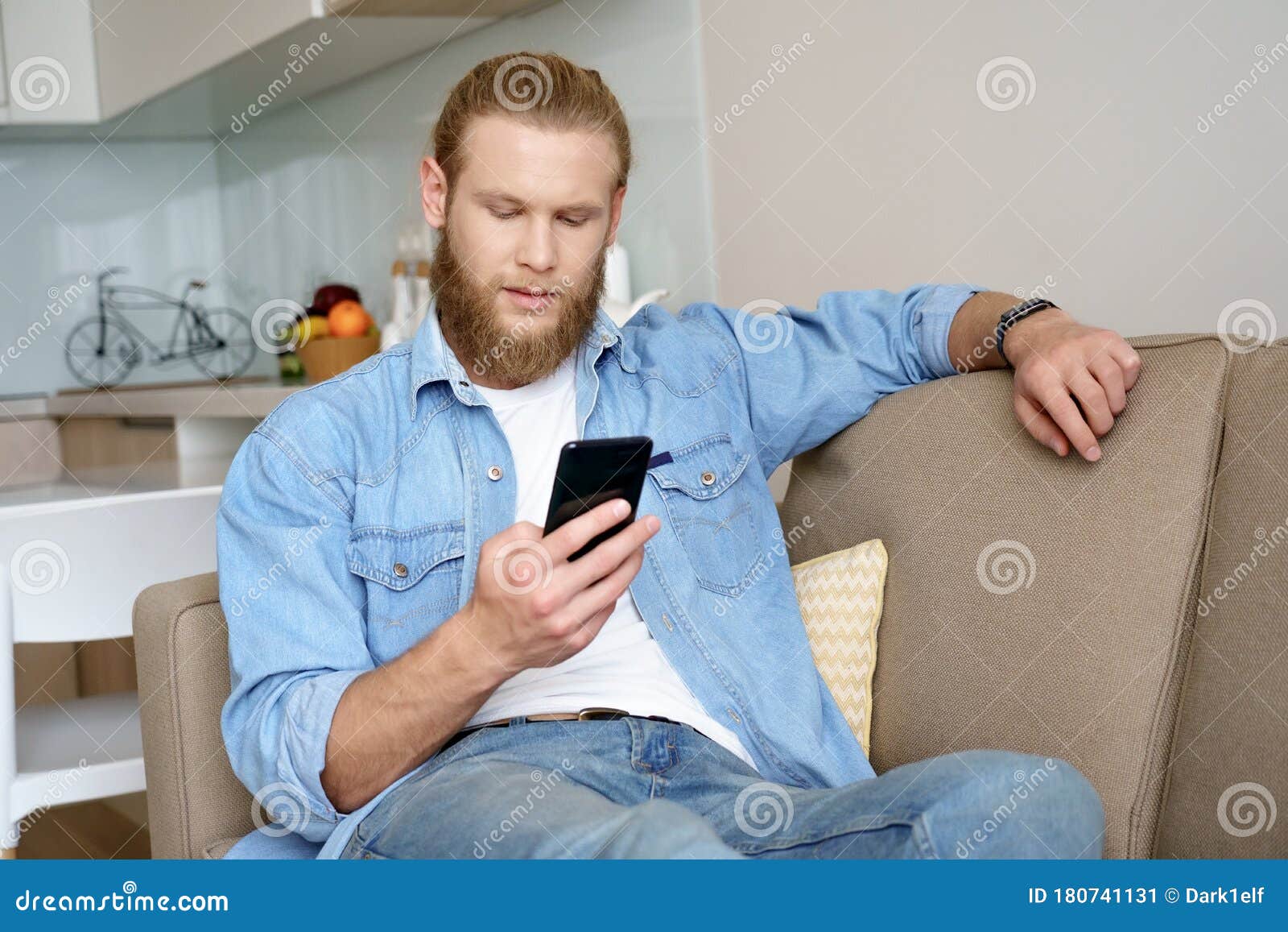 Serious Young Bearded Man Holding Smartphone Using Online App On Sofa