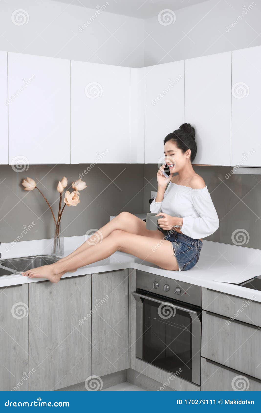 Asian Woman Talking By Mobile Phone While Sitting In The Kitchen Stock