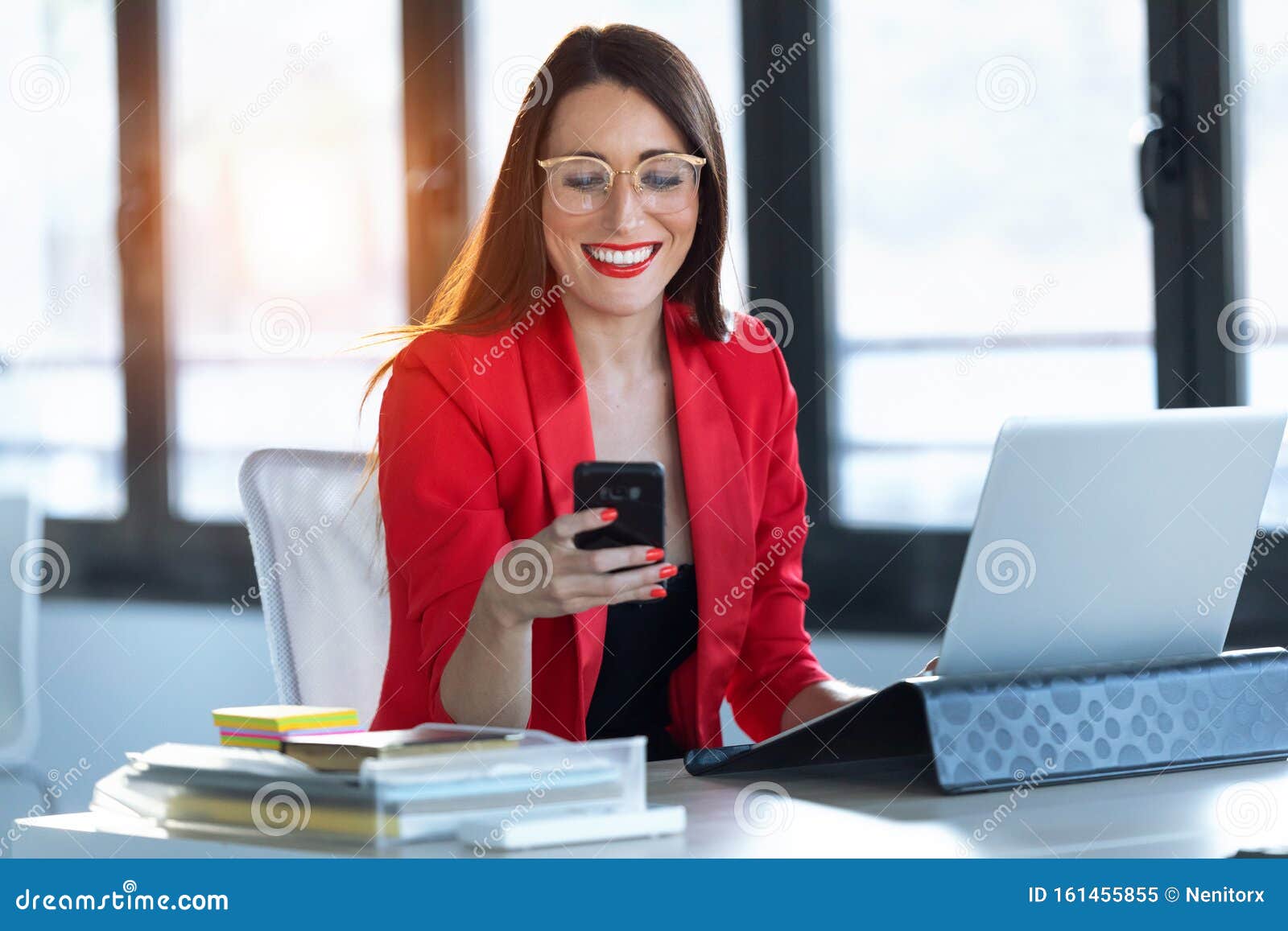 Smiling Young Businesswoman Using Her Mobile Phone while Working with ... Smiling Young Businesswoman Using Her Mobile Phone while Working with ...