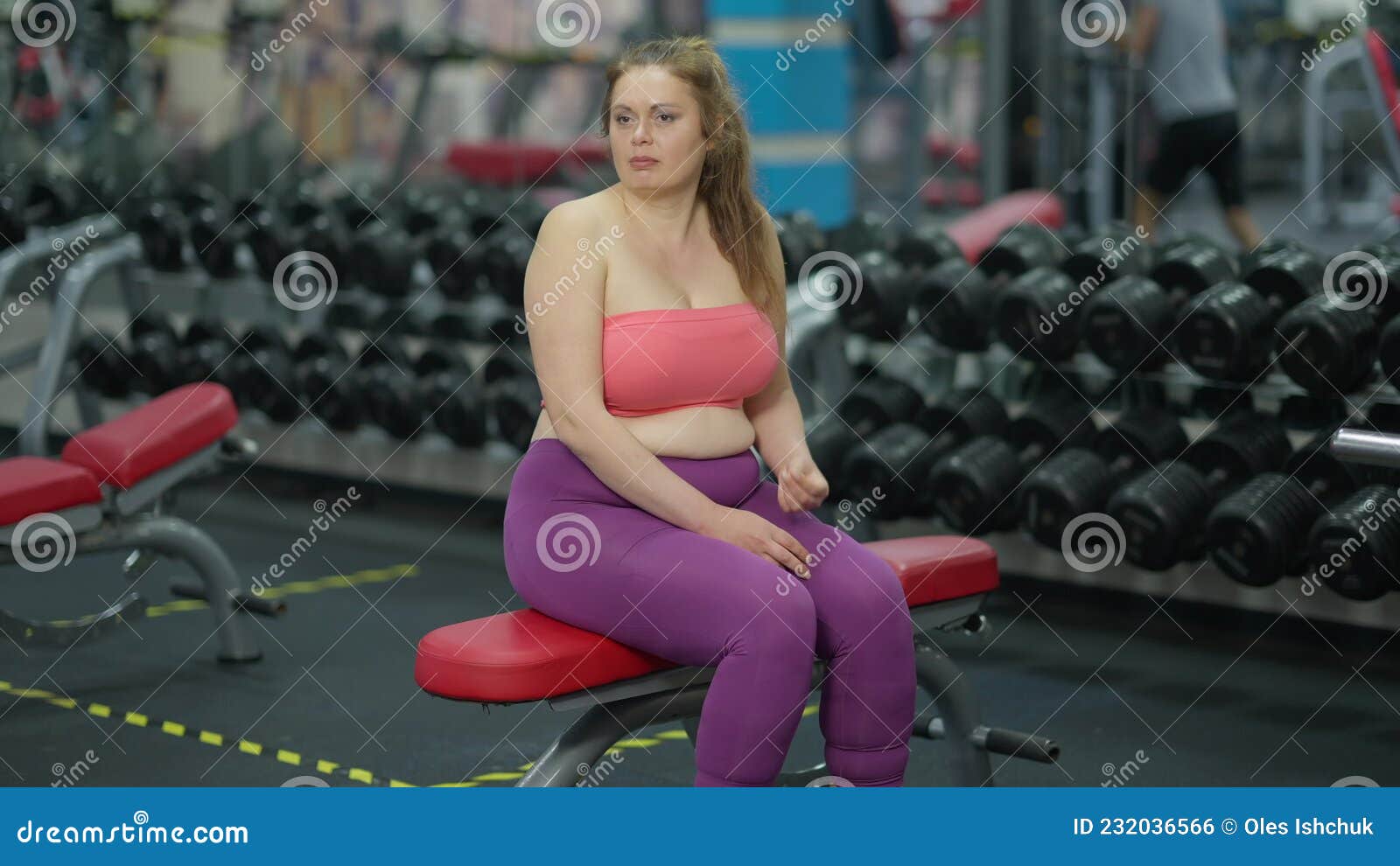 Shy Overweight Woman Sitting In Gym On Exercise Bench Looking Around