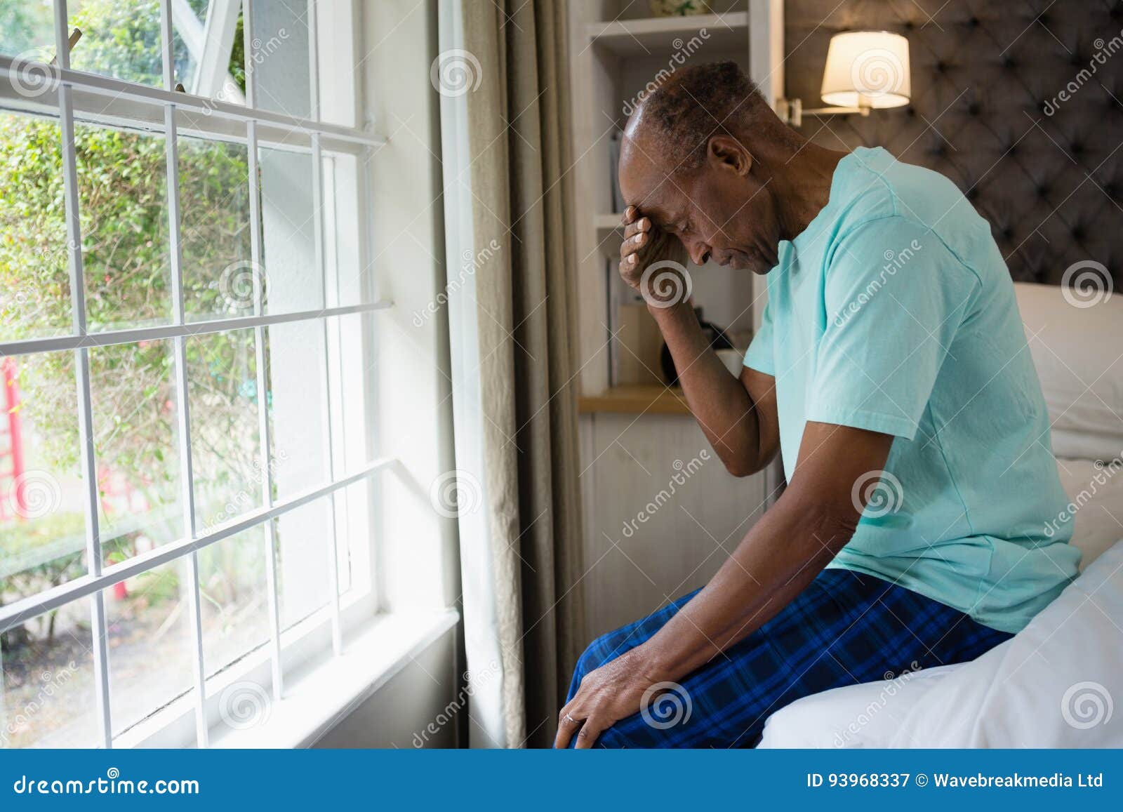 Side View Of Sad Senior Man Sitting On Bed By Window At Home Stock