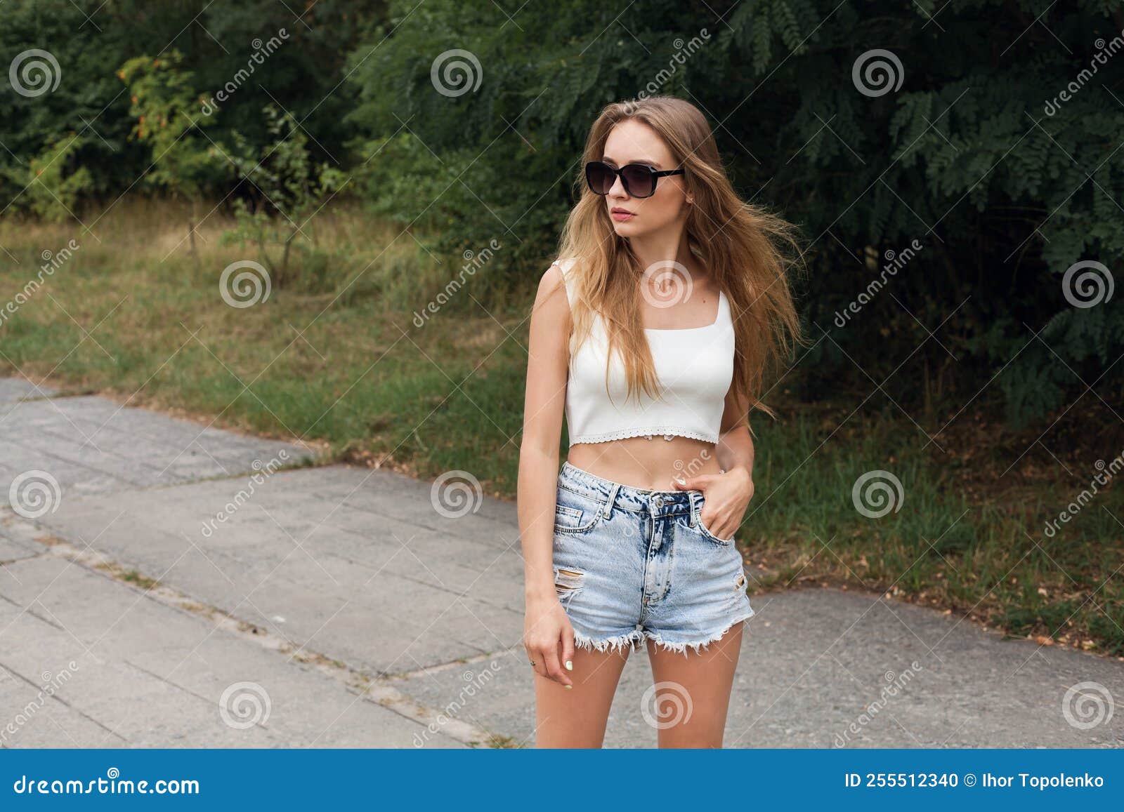 Slender Girl With Long Blond Hair In Short Denim Shorts And A White Top In Sunglasses Stands
